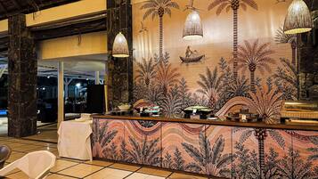 Uma área de recepção com parede decorativa mostrando palmeiras e um pescador, balcão com luminária de palha e piso de azulejos.