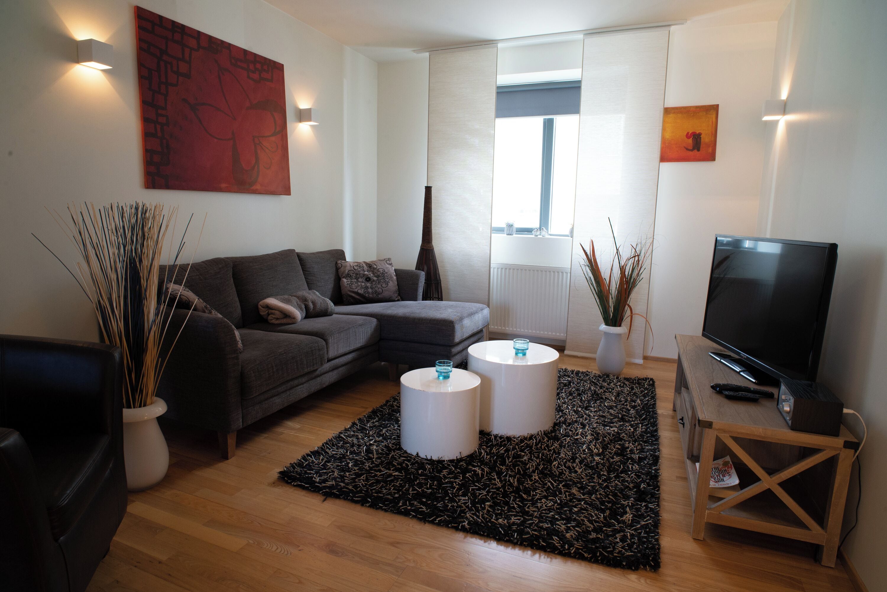 Uma sala de estar moderna com um sofá, duas mesas laterais brancas, uma televisão e uma janela grande.