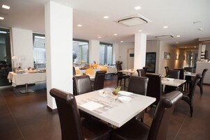 Um restaurante moderno com mesas brancas e cadeiras escuras, janelas amplas e ambiente iluminado.