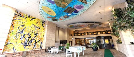 Um hall amplo com teto alto, um mural grande na parede esquerda e uma área de restaurante com mesas ao ar livre.
