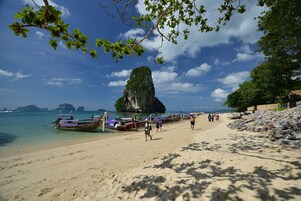 Uma praia com barcos, pessoas e uma formação rochosa imponente.