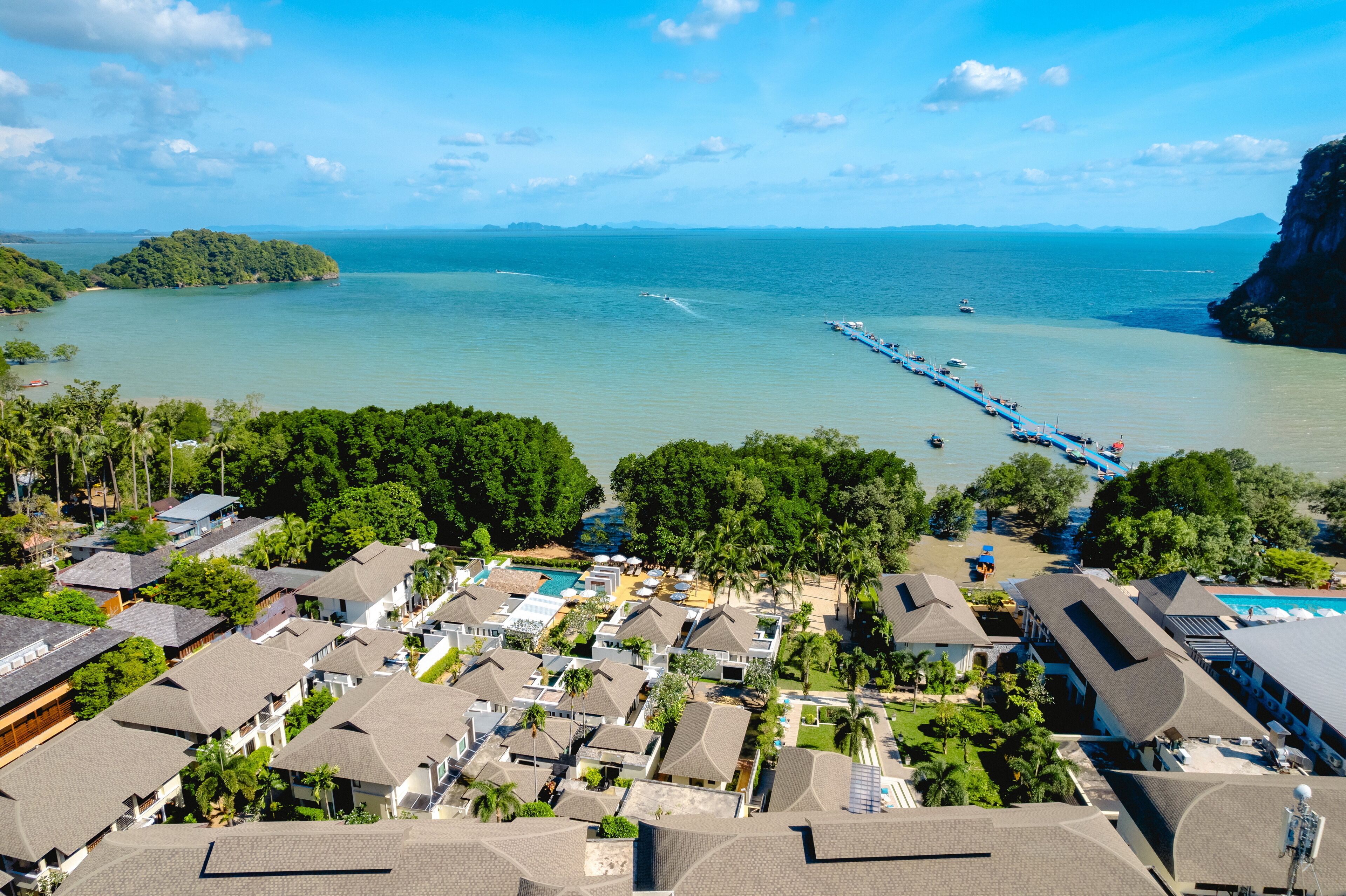 Um resort costeiro com vários edifícios no estilo de bangalôs, um píer que se estende para o mar e vegetação exuberante.