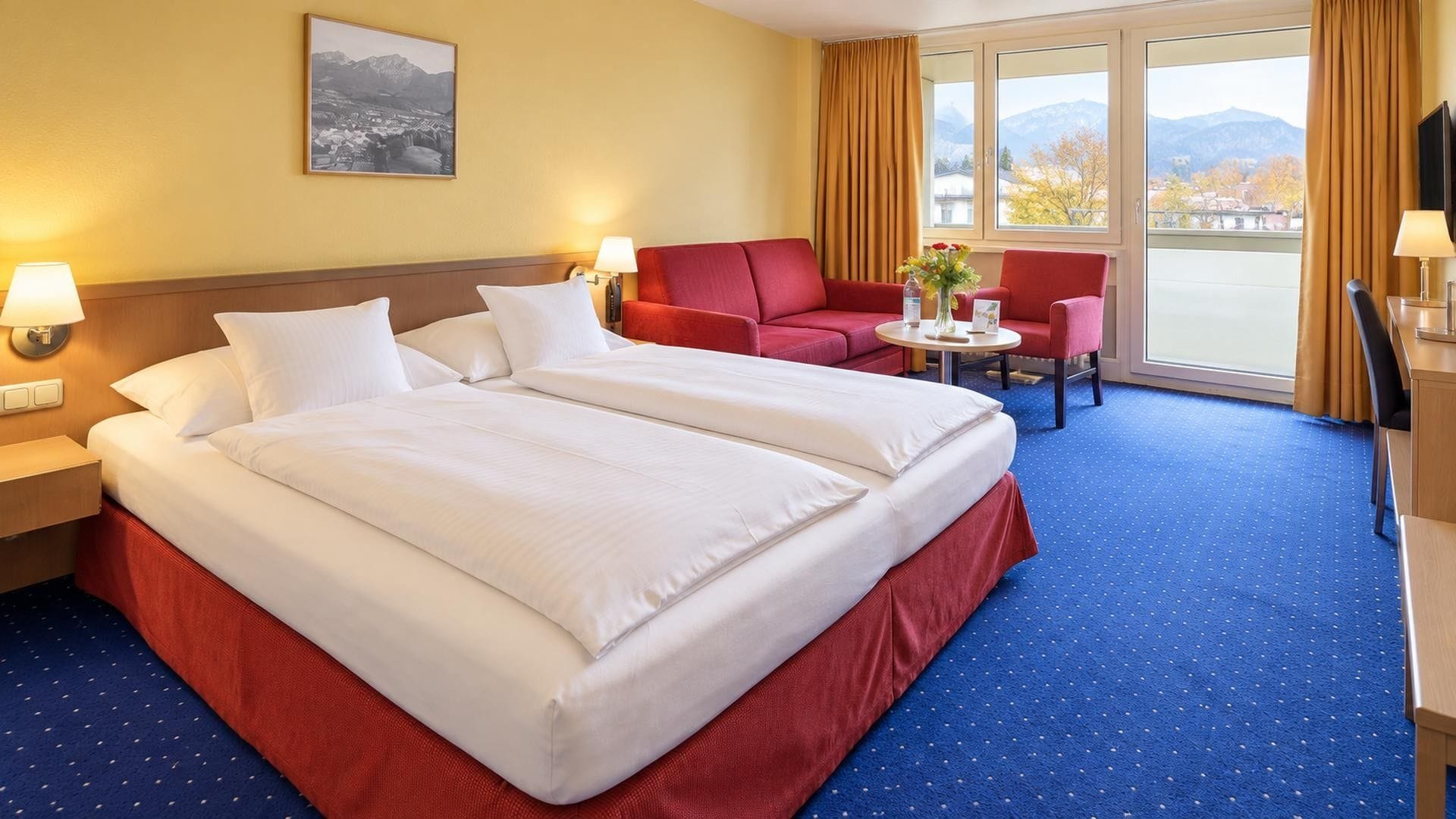 Quarto de hotel com cama de casal, sofá vermelho, mesa pequena com um vaso e televisão na parede.