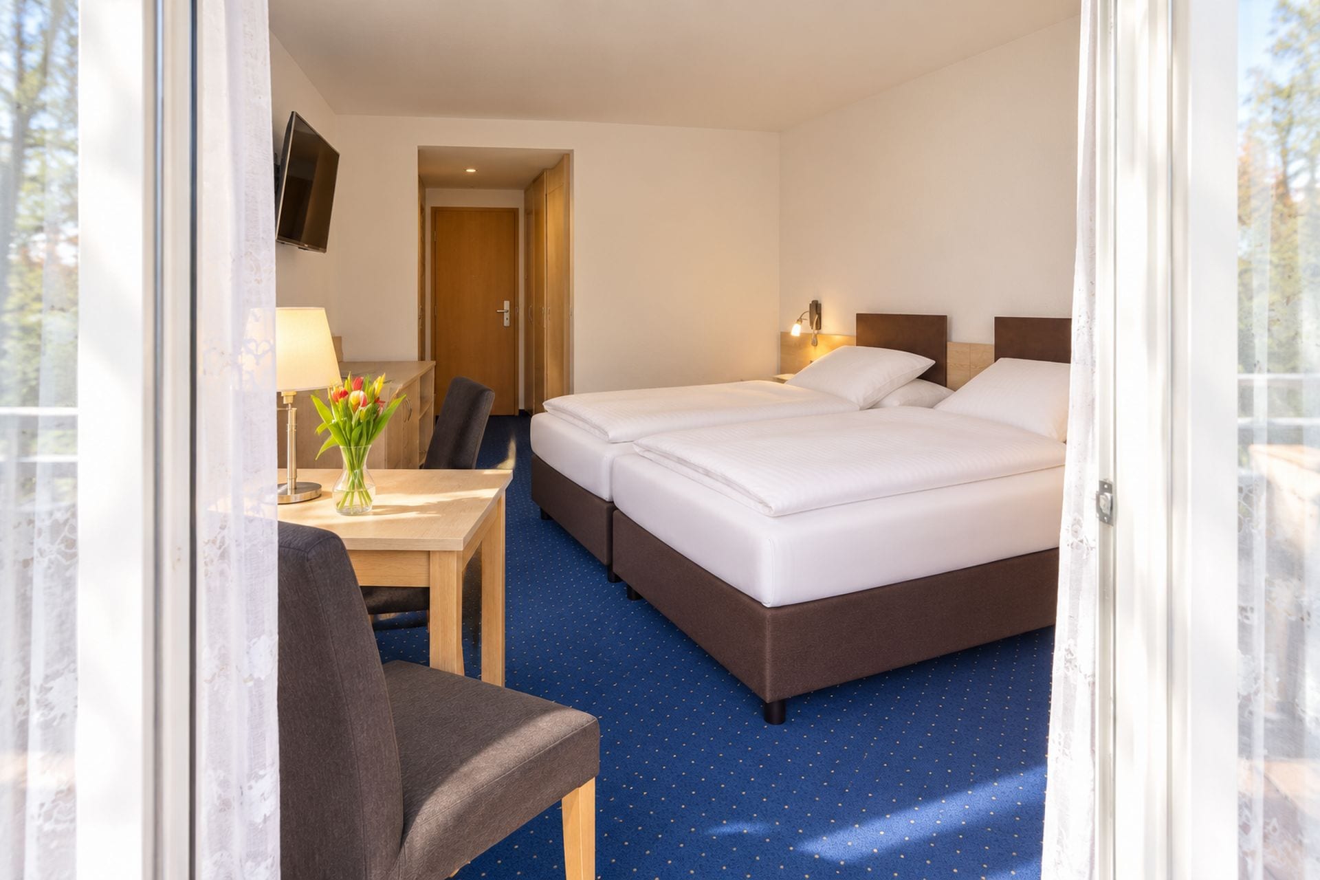 Quarto de hotel com duas camas, uma mesa com luminária e uma cadeira.