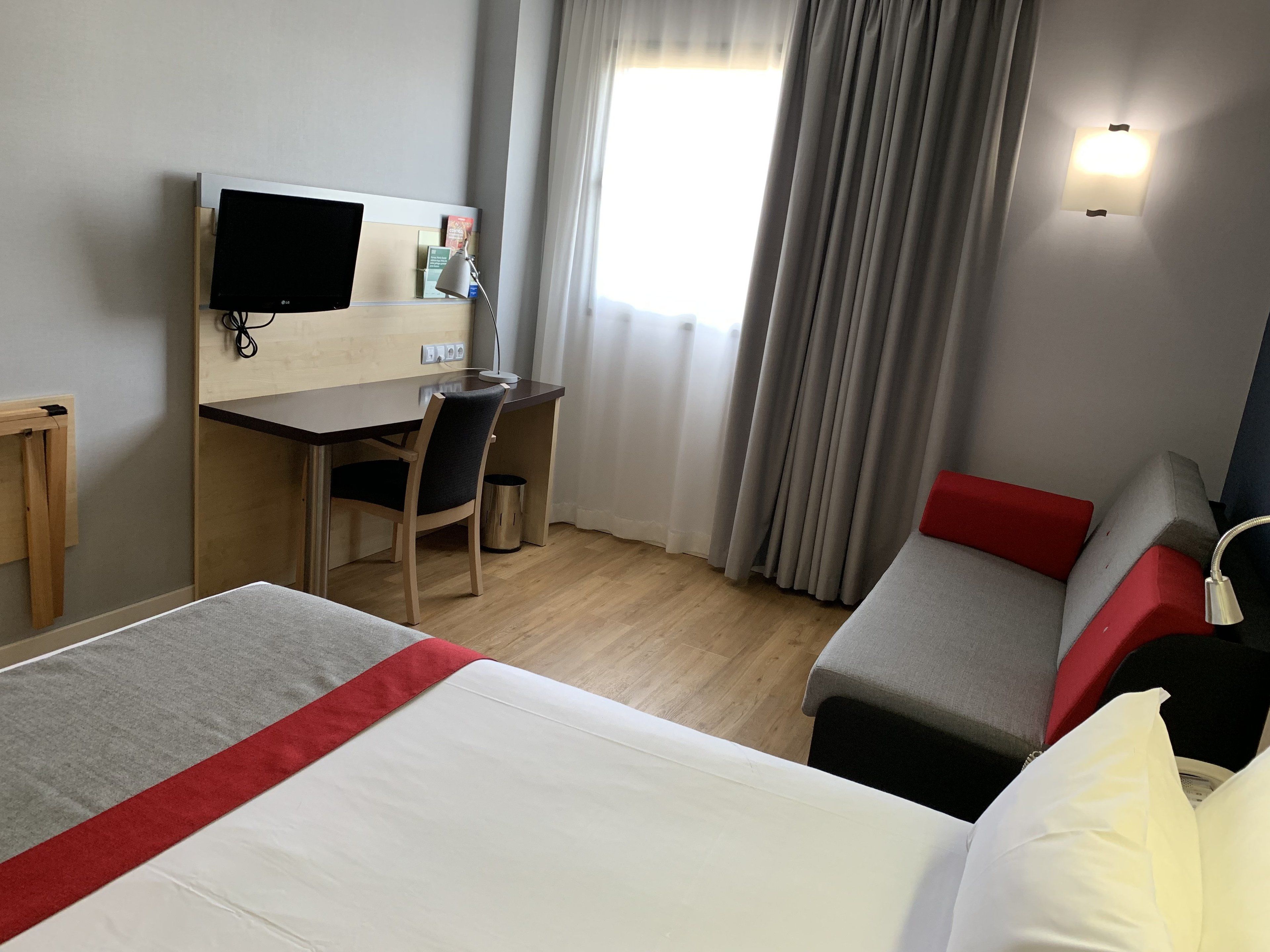Quarto de hotel com cama, escrivaninha, cadeira, televisão e janela com cortinas.