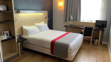 Quarto de hotel com cama, mesa de cabeceira, escrivaninha, cadeira e televisão.