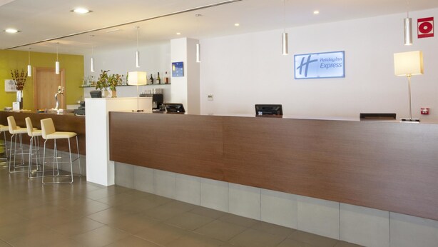 Uma área de recepção com um balcão de madeira, vários bancos altos, uma planta em vaso e uma placa da Holiday Inn Express na parede.