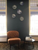 Um quarto com parede escura, uma cadeira marrom, uma mesa redonda pequena com um tabuleiro branco e quatro pratos decorativos na parede.