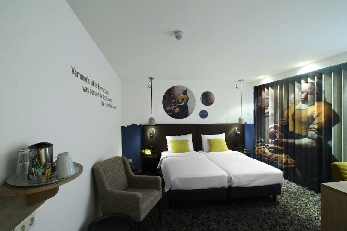 Quarto de hotel com cama de casal, uma cadeira, mesa de cabeceira com abajur e uma obra de arte pendurada na parede.