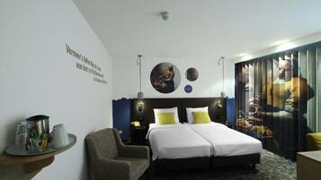 Quarto de hotel com cama de casal, uma cadeira, mesa de cabeceira com abajur e uma obra de arte pendurada na parede.