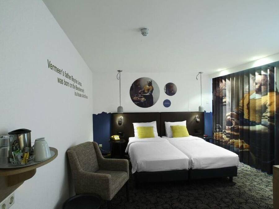Quarto de hotel com cama de casal, uma cadeira, mesa de cabeceira com abajur e uma obra de arte pendurada na parede.