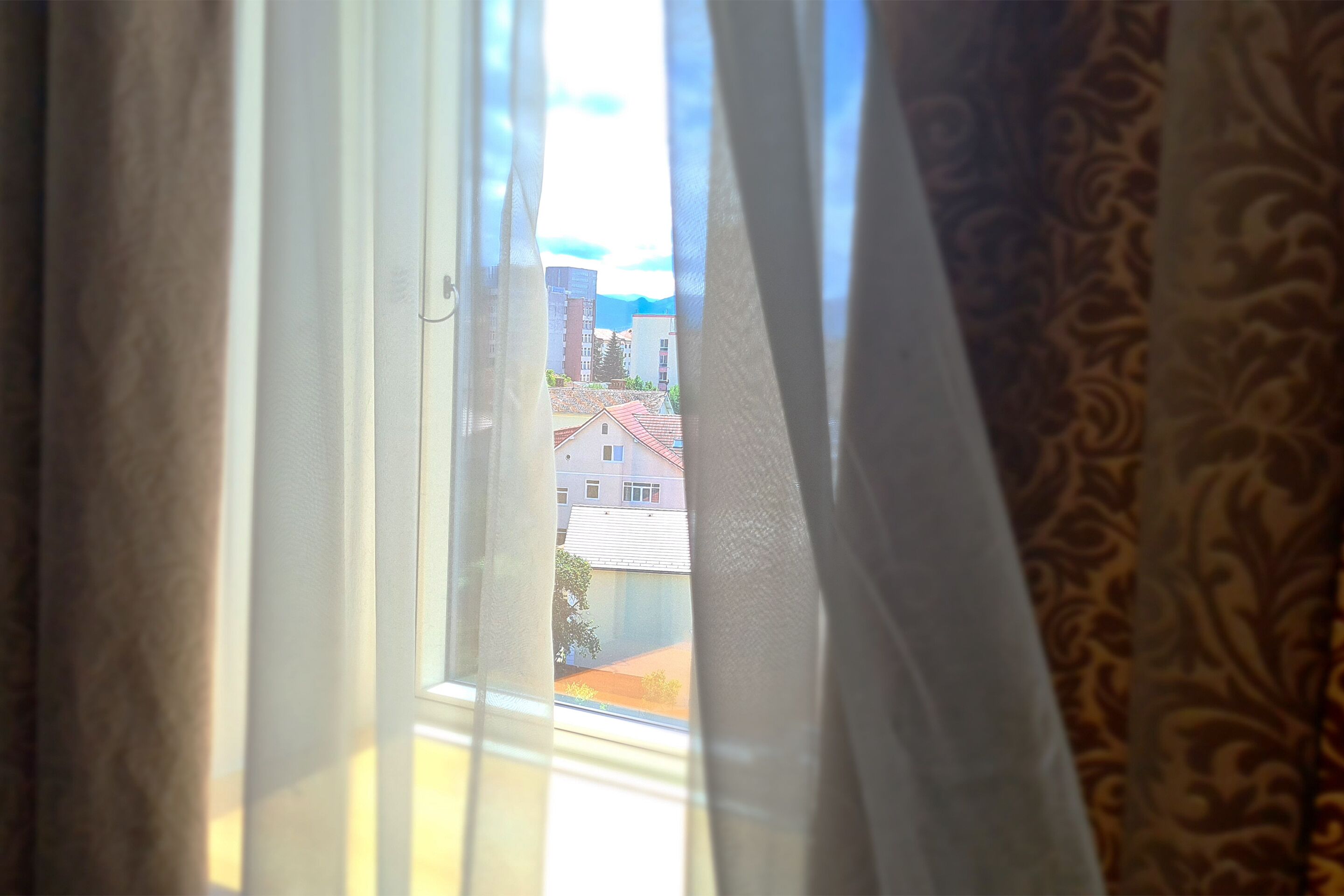 Janela com cortinas leves, mostrando vista de edifícios e uma montanha ao longe.