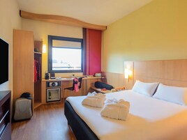 Quarto de hotel com cama, escrivaninha, cadeira, televisão, frigobar e janela com vista.