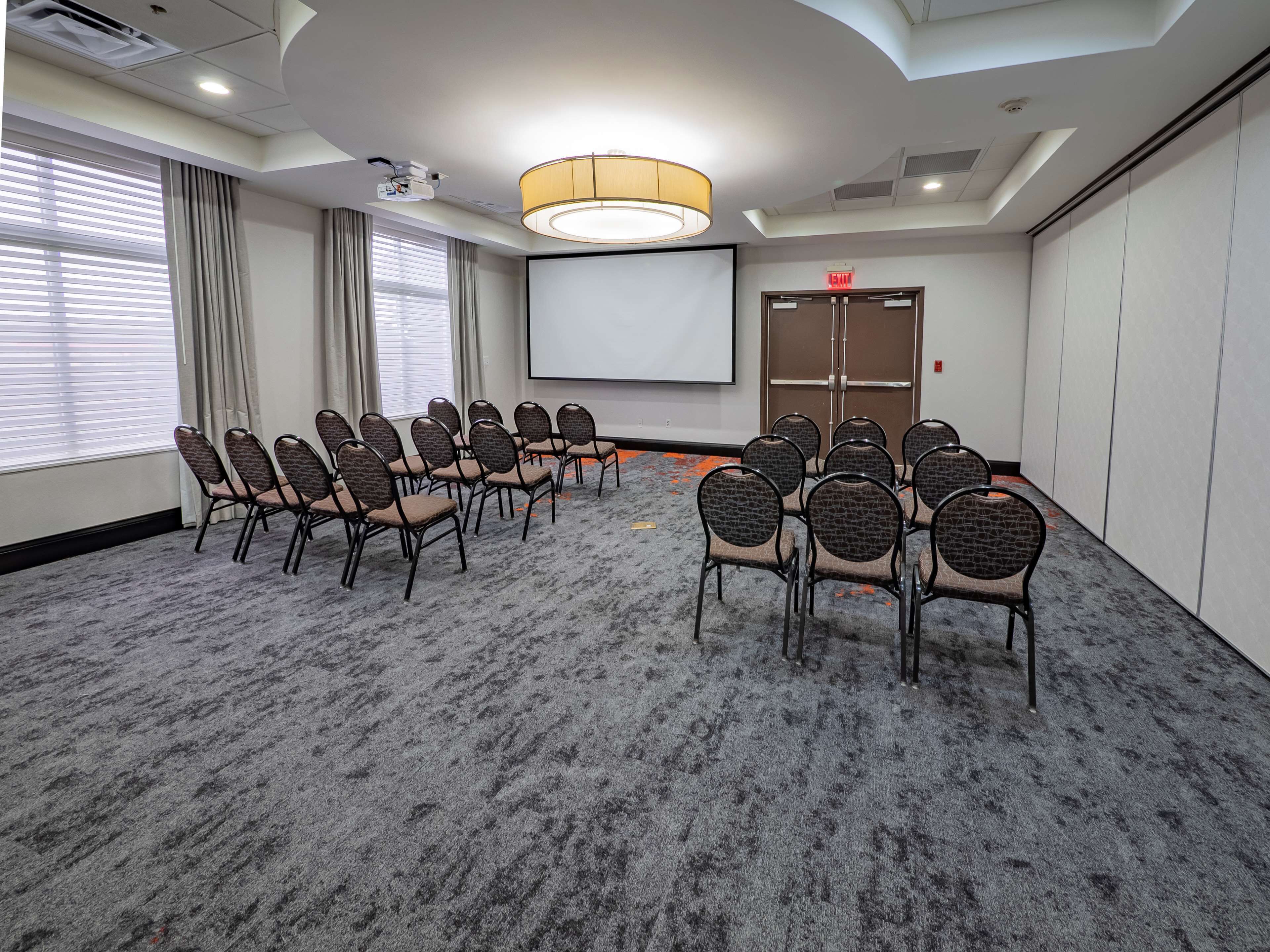 Uma sala de conferências com fileiras de cadeiras, uma tela de projetor e uma janela grande com persianas.