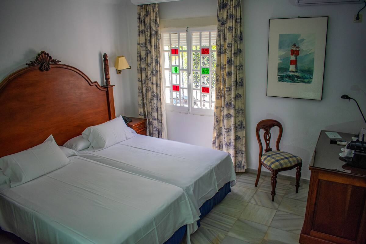 Quarto com duas camas, cabeceira de madeira, uma cadeira, escrivaninha e janela com cortinas.
