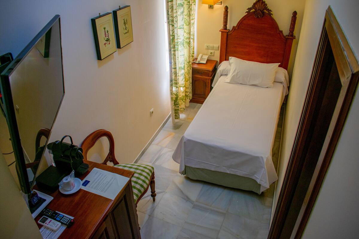 Quarto de hotel com cama, escrivaninha, cadeira, telefone e mesa de cabeceira com abajur.