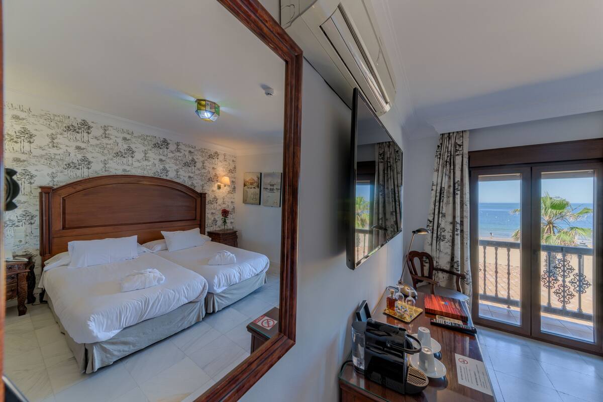 Quarto de hotel com duas camas, um espelho grande, uma televisão, uma mesa de trabalho com telefone e controles remotos, e uma varanda com vista para a praia.