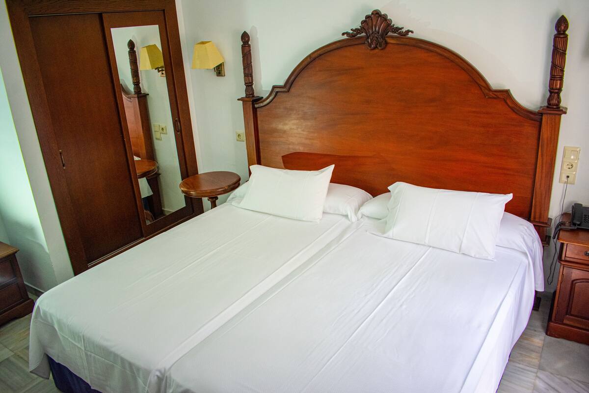 Uma cama de casal com lençóis brancos, uma cabeceira de madeira, uma mesinha de cabeceira e um criado-mudo com abajur.