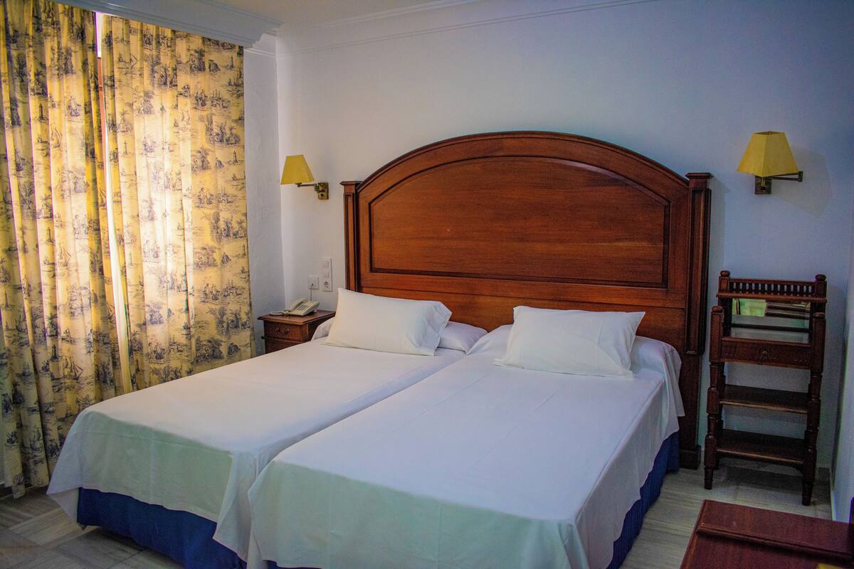 Uma cama de casal com cabeceira de madeira, duas mesas de cabeceira e um criado-mudo com abajur.