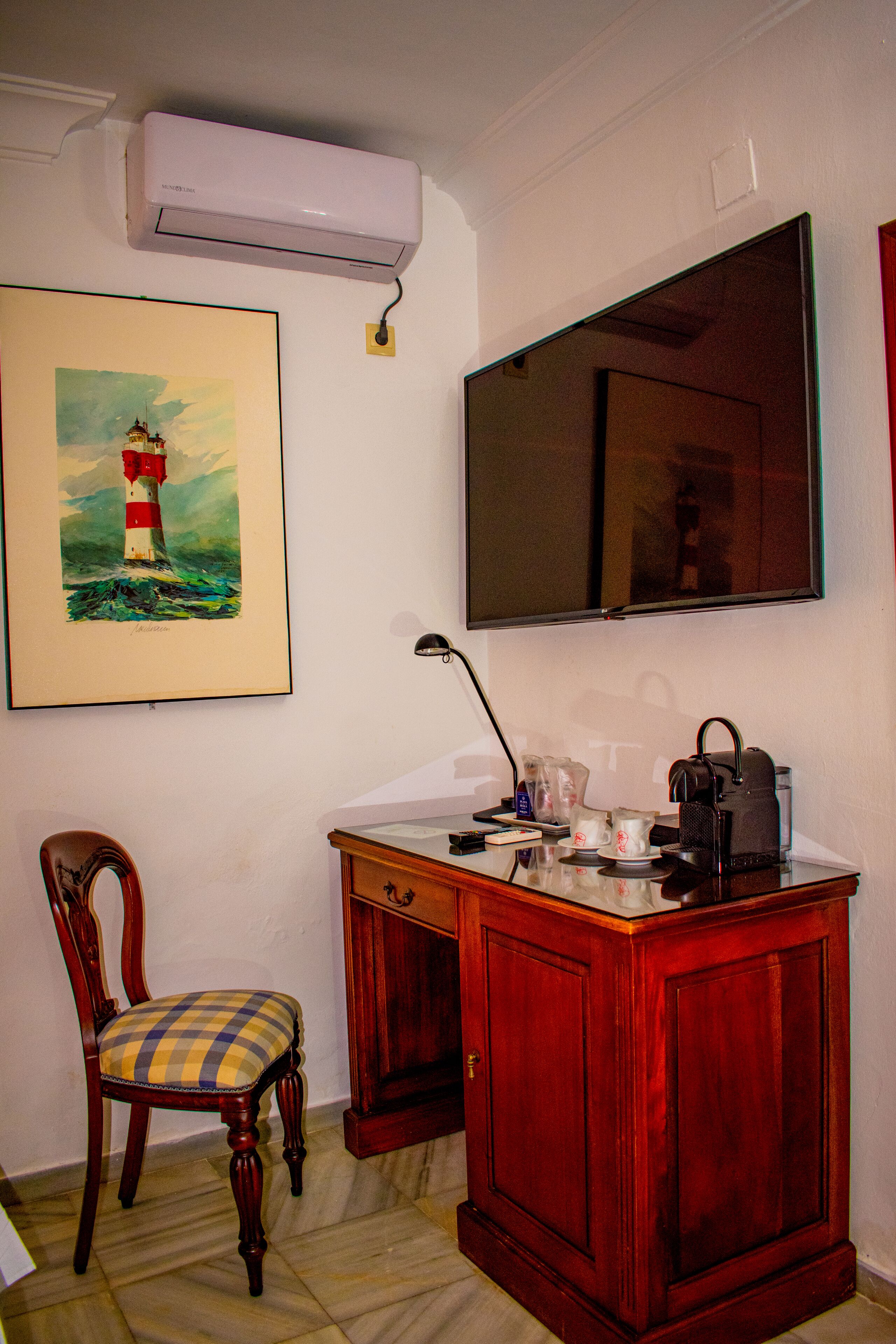 Um quarto com uma escrivaninha de madeira, uma cadeira, um quadro de um farol, uma televisão montada e um aparelho de ar condicionado.