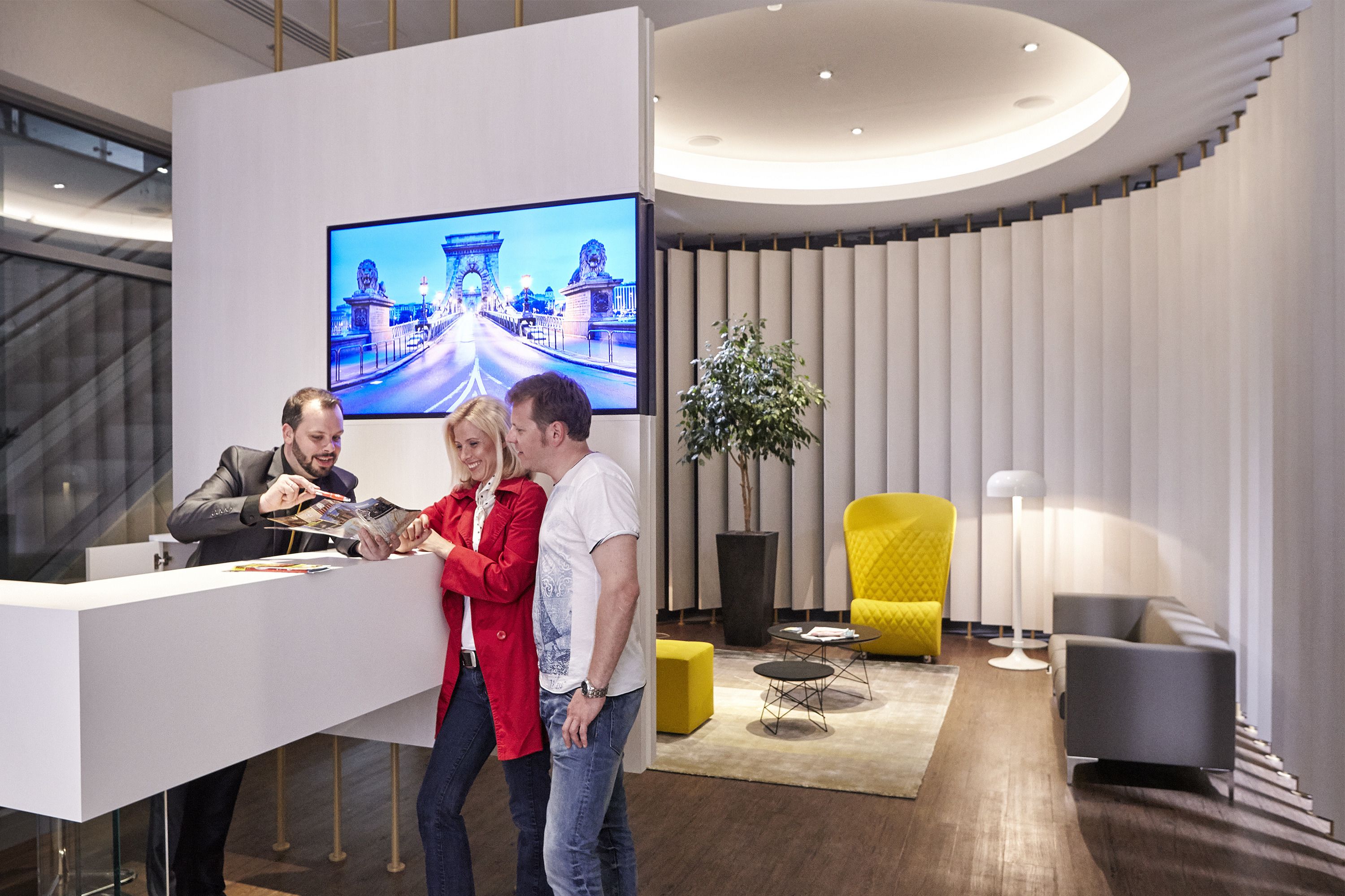 Uma área de recepção com design moderno, apresentando um balcão de atendimento, uma televisão de tela plana grande exibindo uma vista panorâmica, um vaso de planta e assentos contemporâneos, incluindo uma poltrona amarela e um sofá cinza.