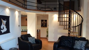 Uma sala de estar ampla com dois sofás de couro preto, piso de madeira, uma escada em espiral e uma cozinha ao fundo.
