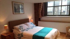 Uma cama bem arrumada com roupa de cama floral, uma mesa de cabeceira com abajur, uma cadeira de vime e uma janela com cortinas.