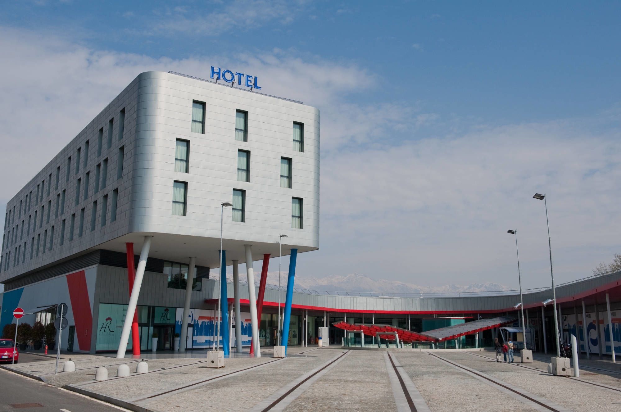 Um hotel moderno com telhado plano, um letreiro chamativo 'HOTEL' e uma área de entrada espaçosa com toldo vermelho e azul.