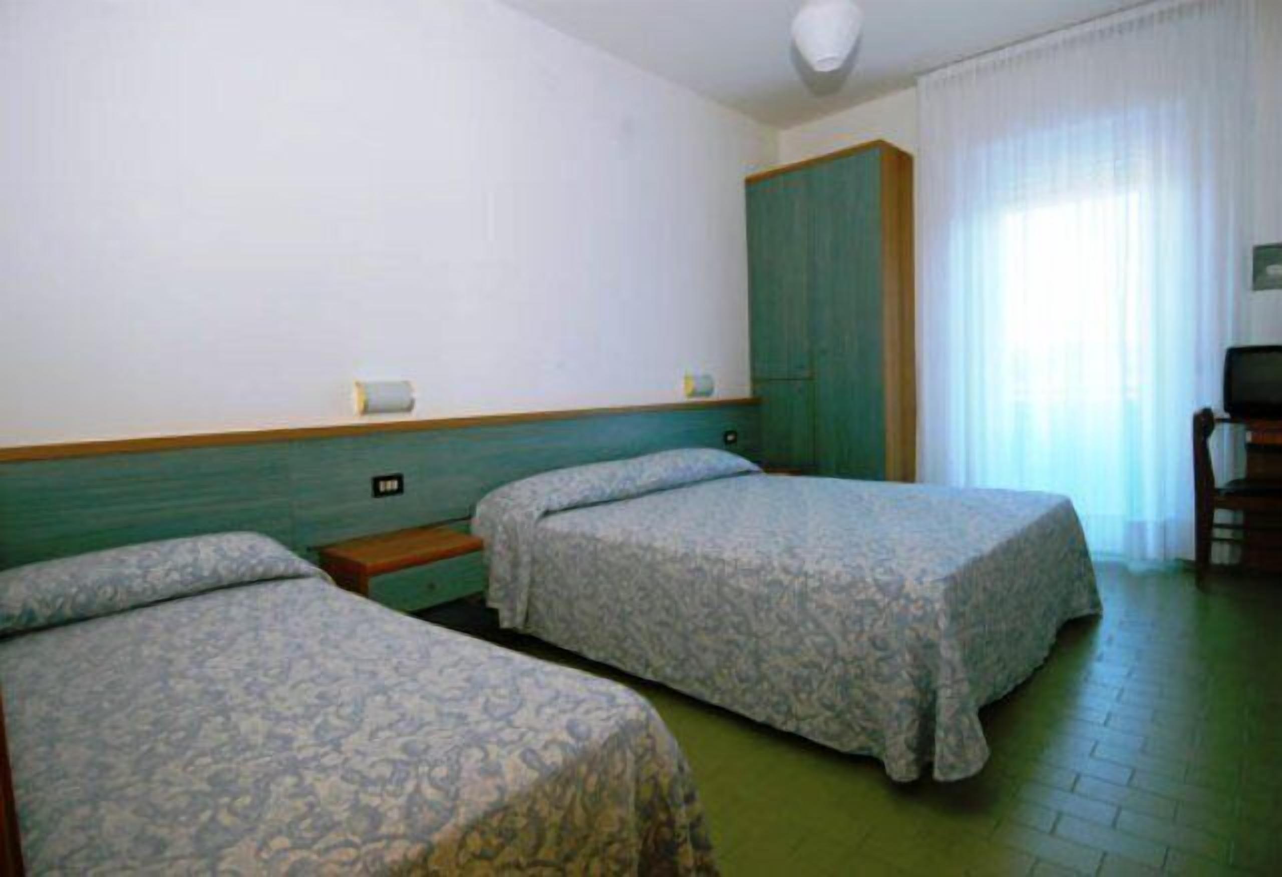 Quarto com duas camas, um banco de madeira, um guarda-roupa verde e uma televisão sobre um móvel de madeira.