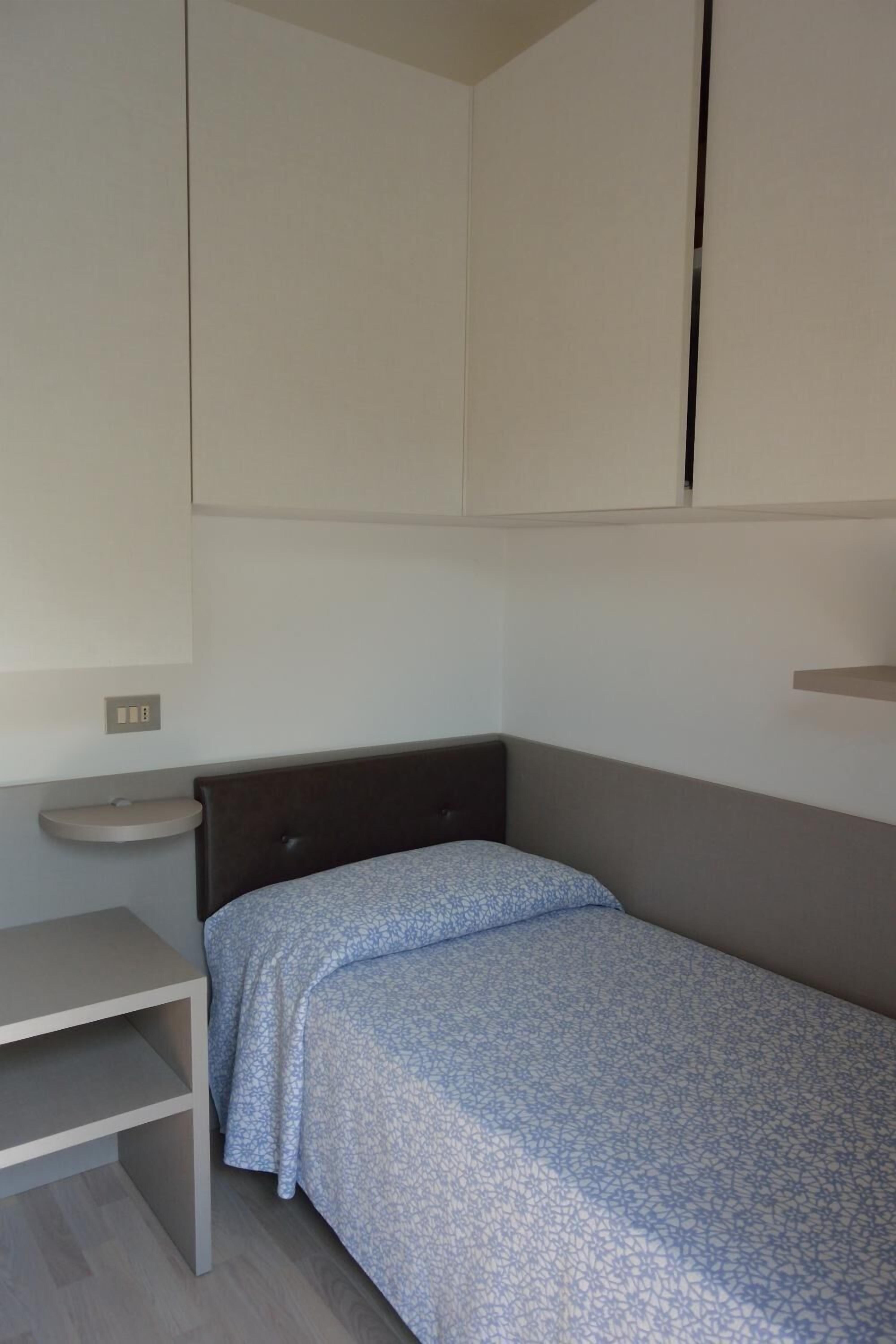 Quarto pequeno com uma cama de solteiro, cabeceira de madeira, mesa de cabeceira e prateleira na parede.