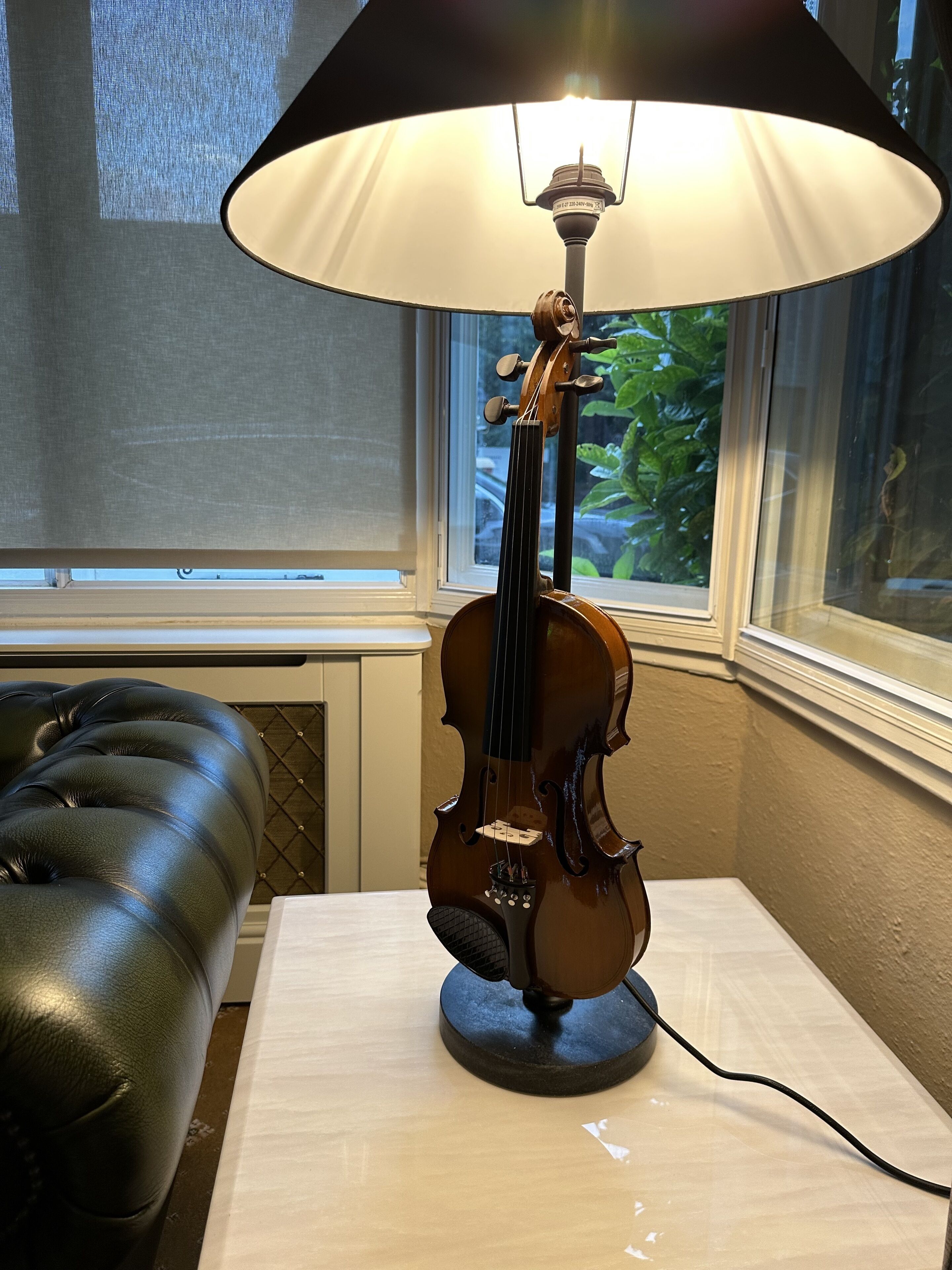 Um violino sob uma lâmpada sobre uma mesa perto de uma janela com vista para vegetação.