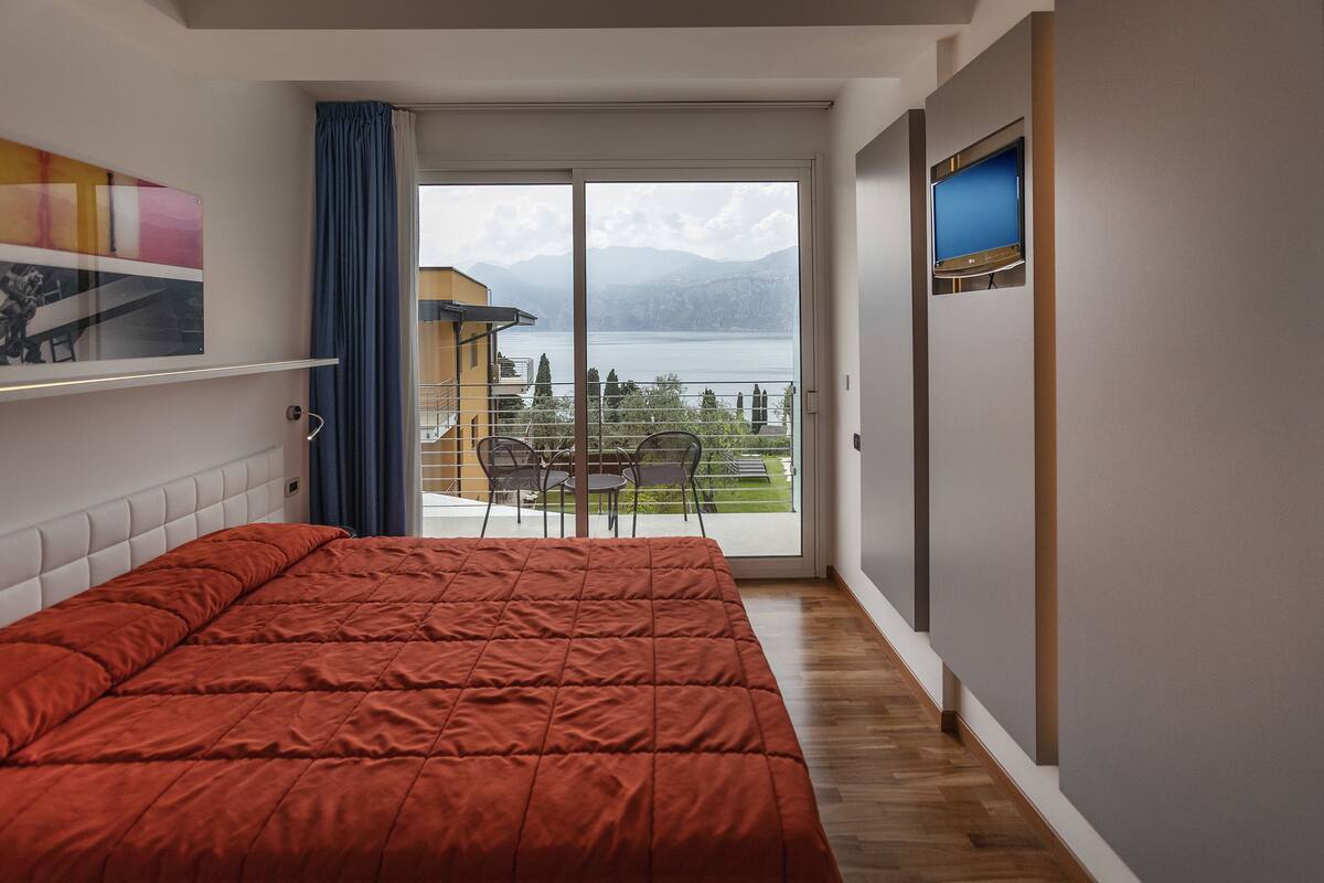 Quarto com cama grande, porta de vidro deslizante para varanda com vista para montanhas e um lago, e televisão na parede.
