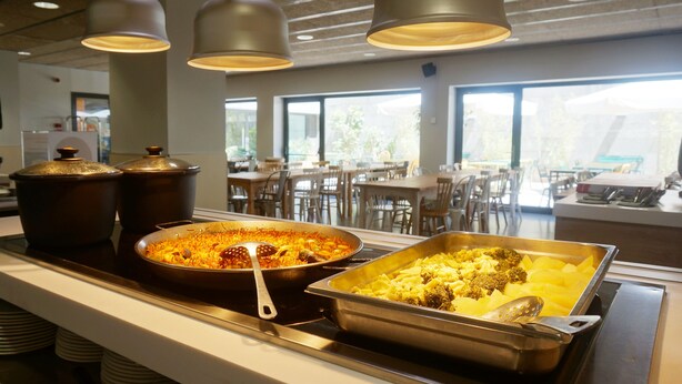 Uma área de buffet com diversos pratos, incluindo massa e vegetais, em um ambiente bem iluminado com amplas janelas e luminárias modernas.