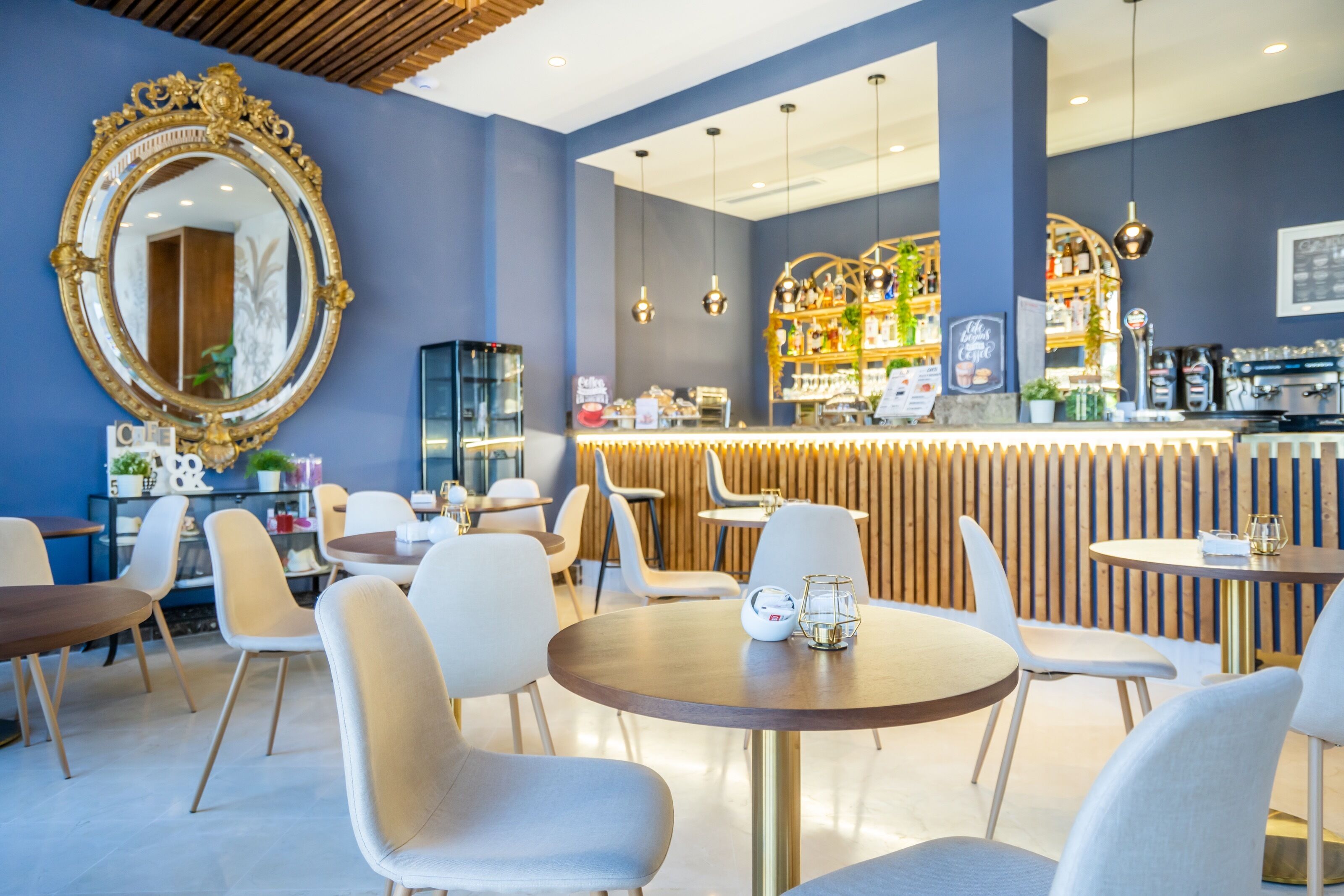 Um café moderno com mesas redondas e cadeiras brancas, uma área de bar com prateleiras de garrafas e um espelho grande e ornamentado na parede.