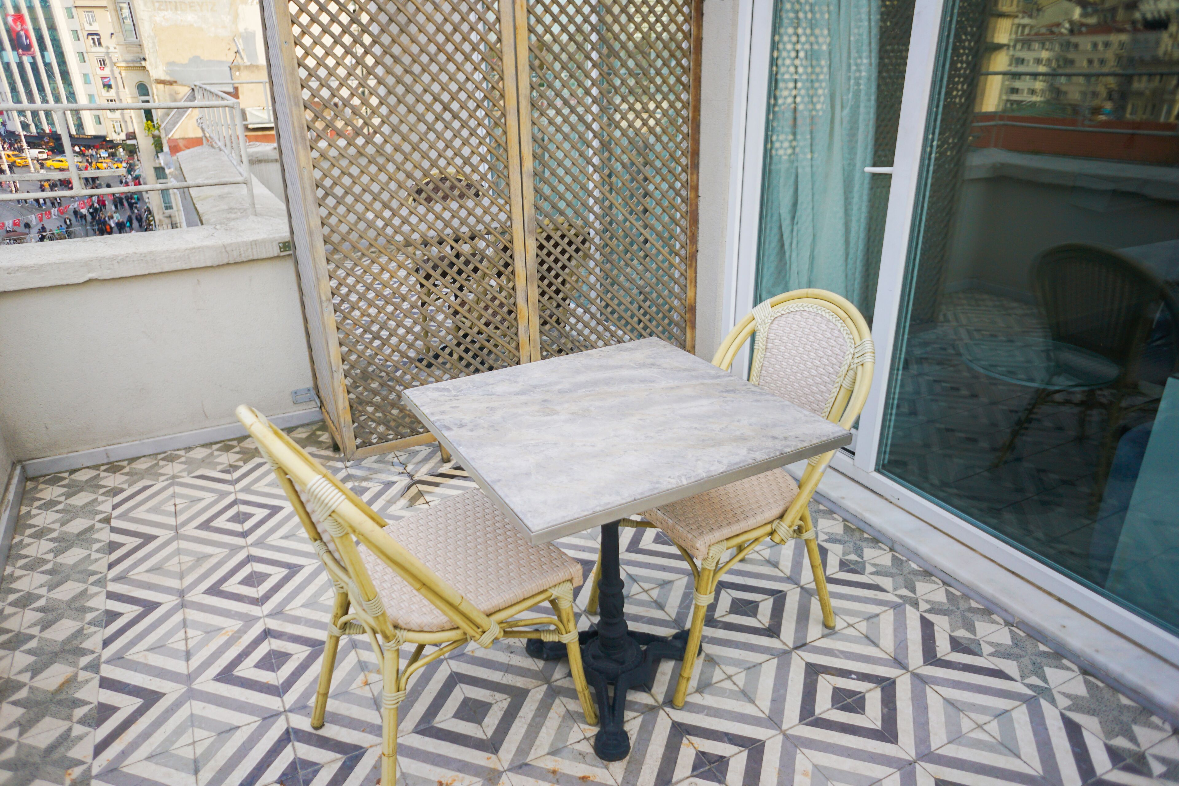 Uma varanda com piso estampado, uma mesa, duas cadeiras e um divisório de vime.