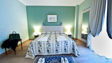 Quarto de hotel com uma cama, duas luminárias de cabeceira, uma parede verde, um quadro emoldurado, uma bolsa de mão verde e um par de chinelos sobre um tapete azul.
