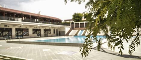 Uma área de piscina com um deck ladrilhado, um espaço coberto para sentar e um edifício com telhado vermelho.