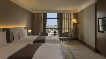 Quarto de hotel com duas camas, uma cadeira, televisão e vista para a cidade.