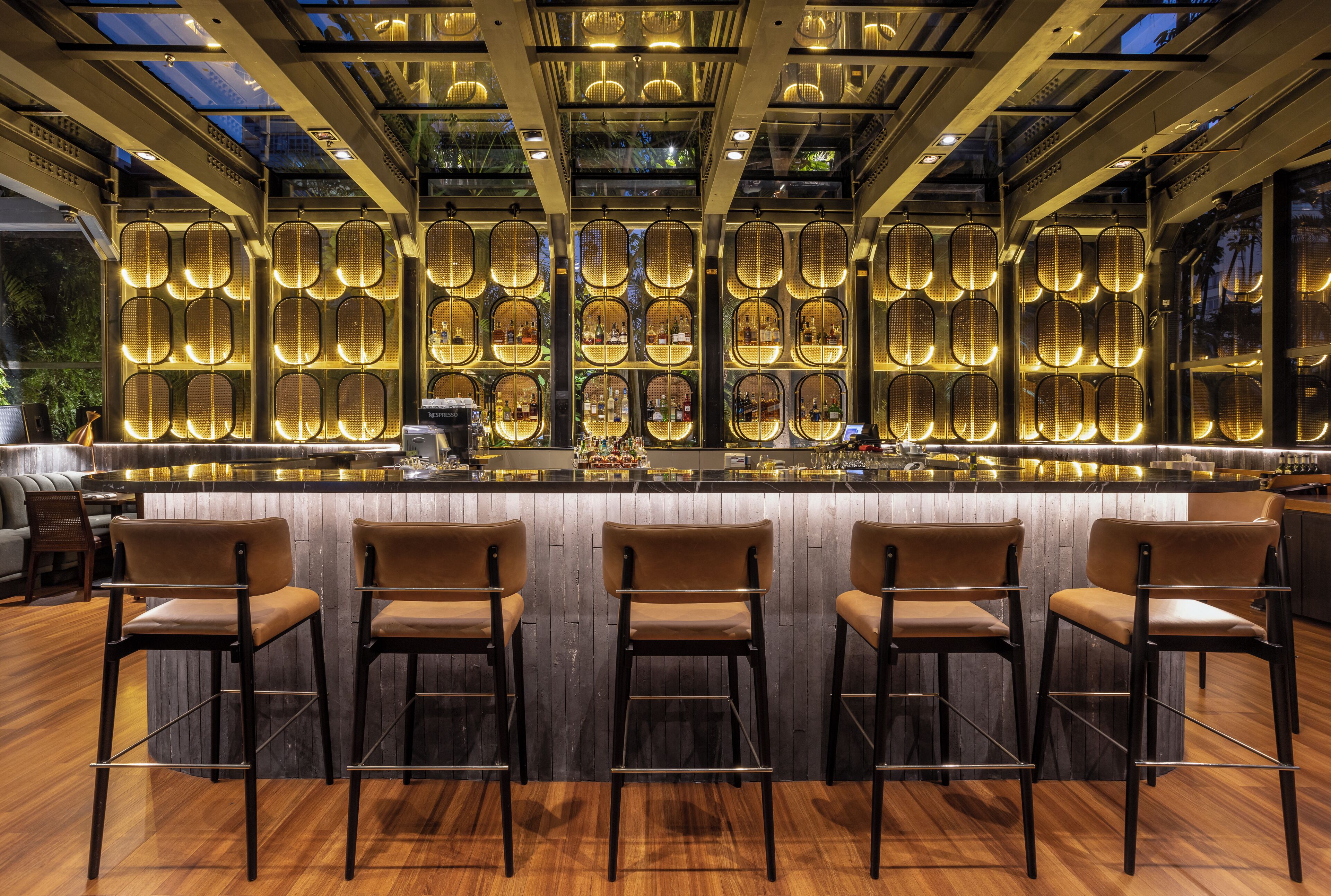 Uma área de bar com piso de madeira, banquetas e uma parede de prateleiras iluminadas.