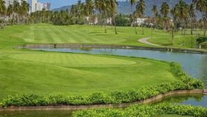 Um campo de golfe com um lago, palmeiras e montanhas ao fundo.