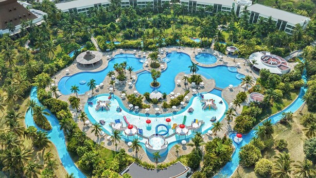 Vista aérea de um resort com várias piscinas, palmeiras e um elemento central de água.