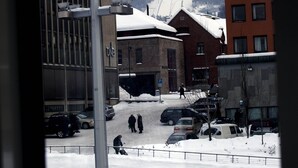 Rua nevada em área urbana, com edifícios, carros estacionados e uma pessoa caminhando.
