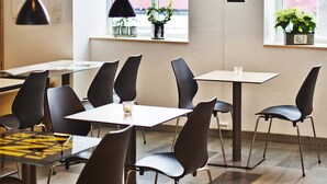 Interior de café moderno com cadeiras pretas, mesas brancas, luminárias pendentes e plantas em vasos.