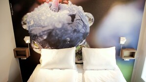 Cama com lençóis brancos e duas almofadas, mesa de cabeceira fixada na parede com abajur, e pôster de um objeto azul com gotas de água.