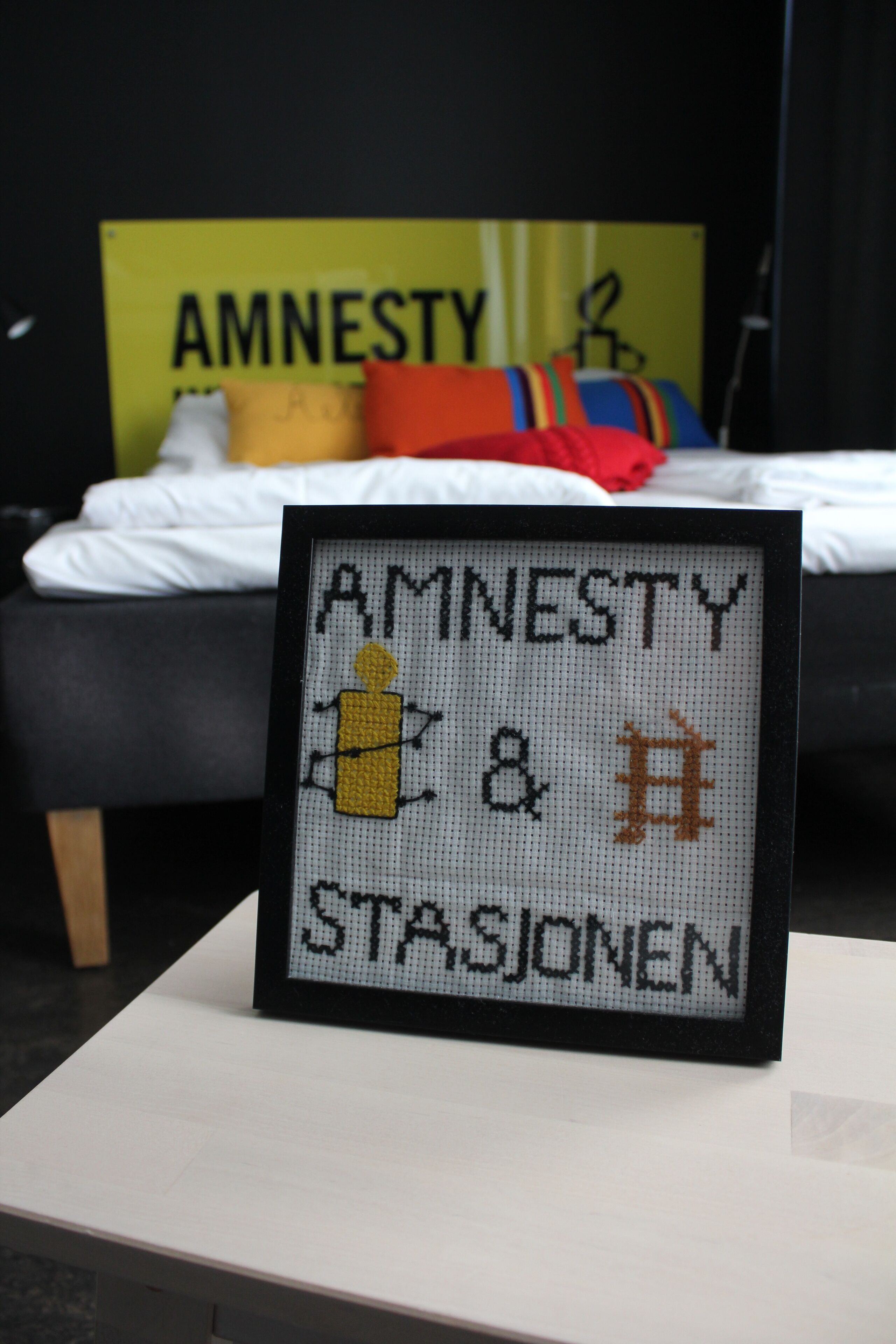 Uma obra de bordado em moldura com as palavras "AMNESTY" e "STATIONEN" sobre uma mesa branca.