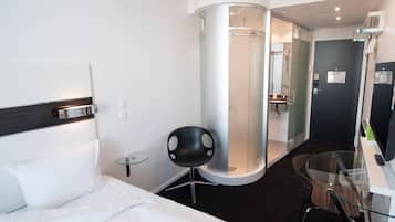 Quarto de hotel moderno com cama, chuveiro em vidro, uma pequena mesa e uma cadeira.