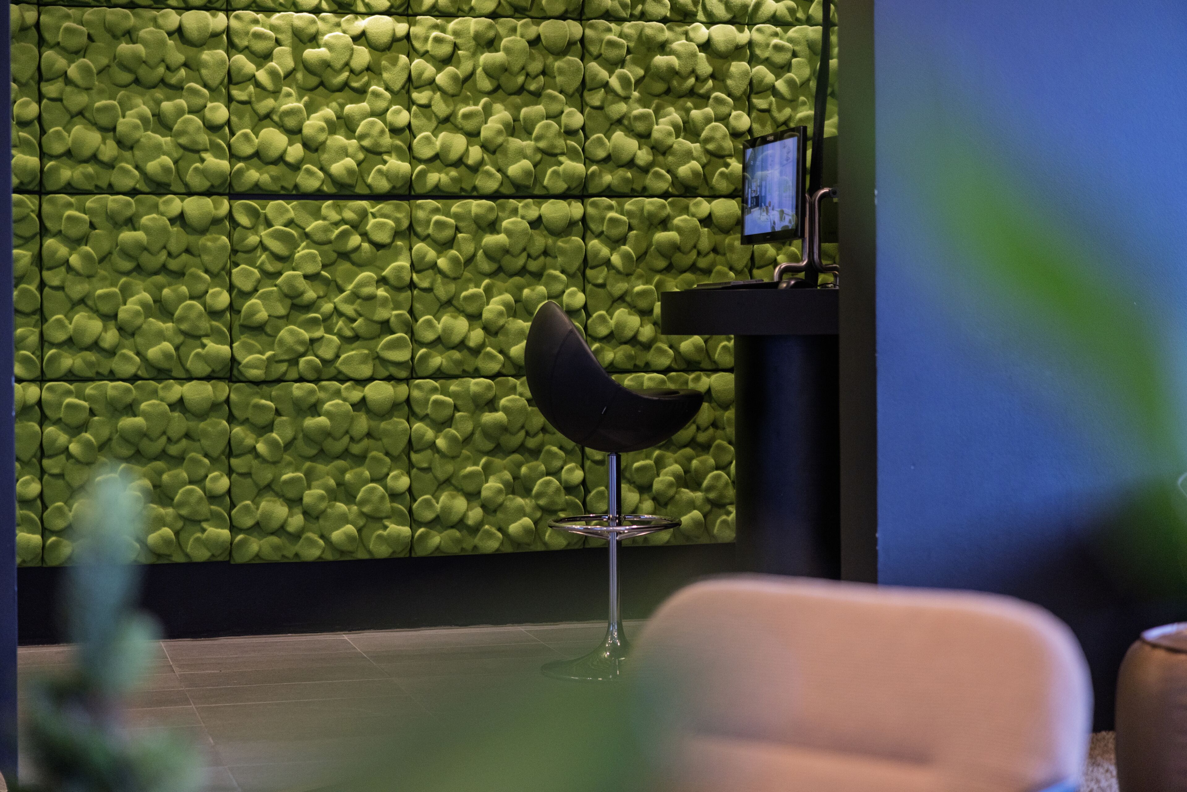 Uma área de recepção com parede verde texturizada, uma cadeira preta moderna e uma pequena mesa com uma tela.