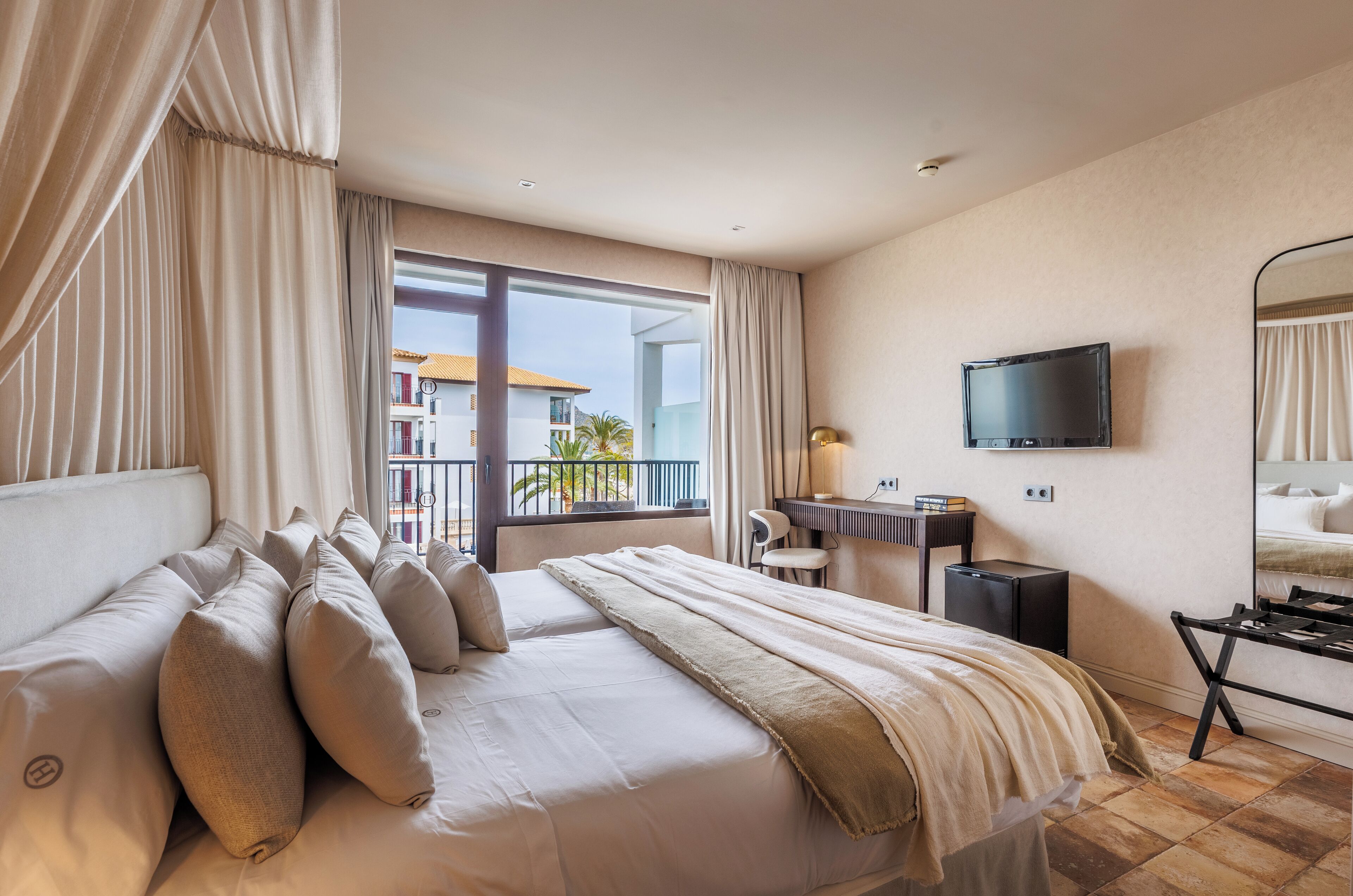 Quarto de hotel com uma cama grande, uma mesa, uma cadeira, uma TV e uma varanda com vista para edifícios e palmeiras.