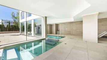 Piscina interior moderna com paredes de vidro transparente, piso azulejado e vista da área circundante.