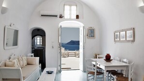 Quarto com decoração clara, área de jantar com mesa e cadeiras, sofá, espelho e vista para o mar através de um vão aberto.
